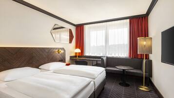 Quarto de hotel com duas camas, uma escrivaninha, um sofá, uma luminária e uma janela grande com cortinas.