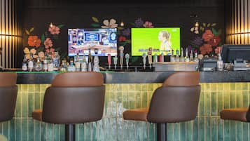Uma área de bar com banquetas, um balcão com garrafas e uma tela de televisão exibindo uma mulher.