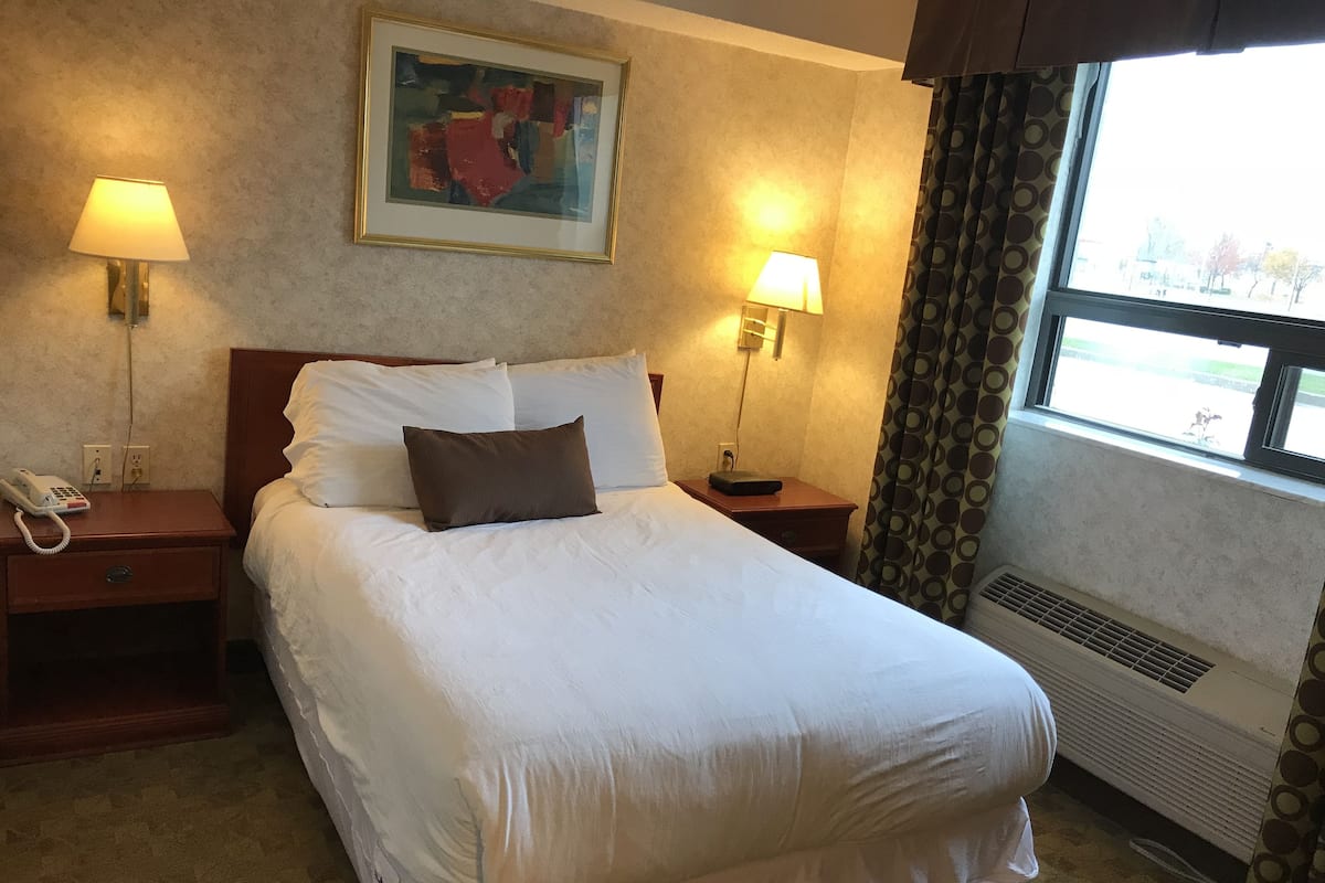 Quarto de hotel com cama arrumada, duas luminárias de cabeceira, um quadro na parede e uma janela com cortinas.