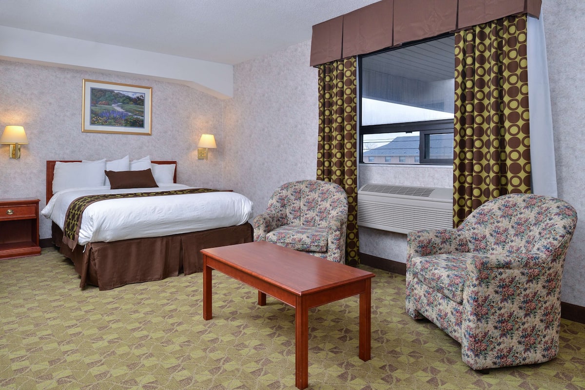 Quarto de hotel com uma cama, duas poltronas, uma mesa de madeira e uma janela grande com cortinas.