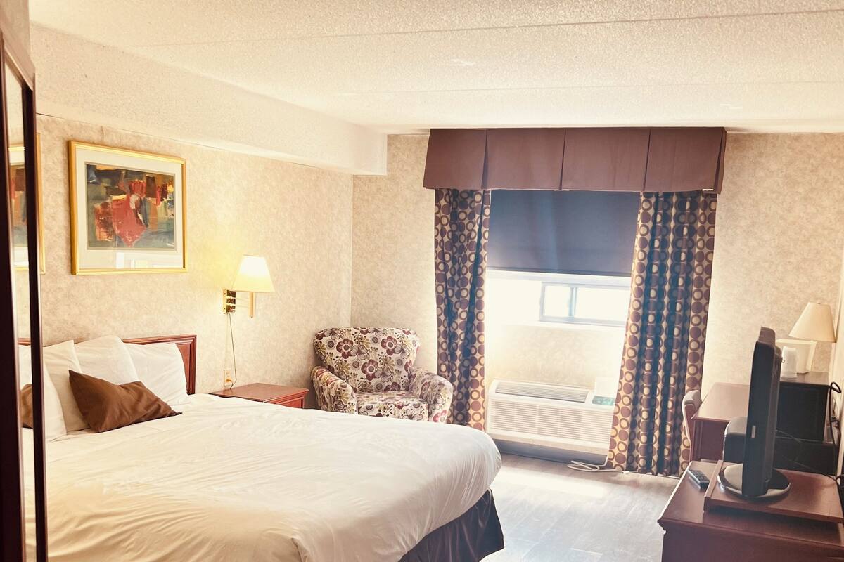 Quarto de hotel com uma cama grande, uma cadeira, uma escrivaninha com computador e uma janela com cortinas.