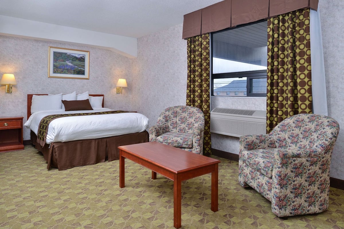 Quarto de hotel com cama, duas poltronas, uma mesa de madeira, um quadro na parede e uma janela com cortinas.
