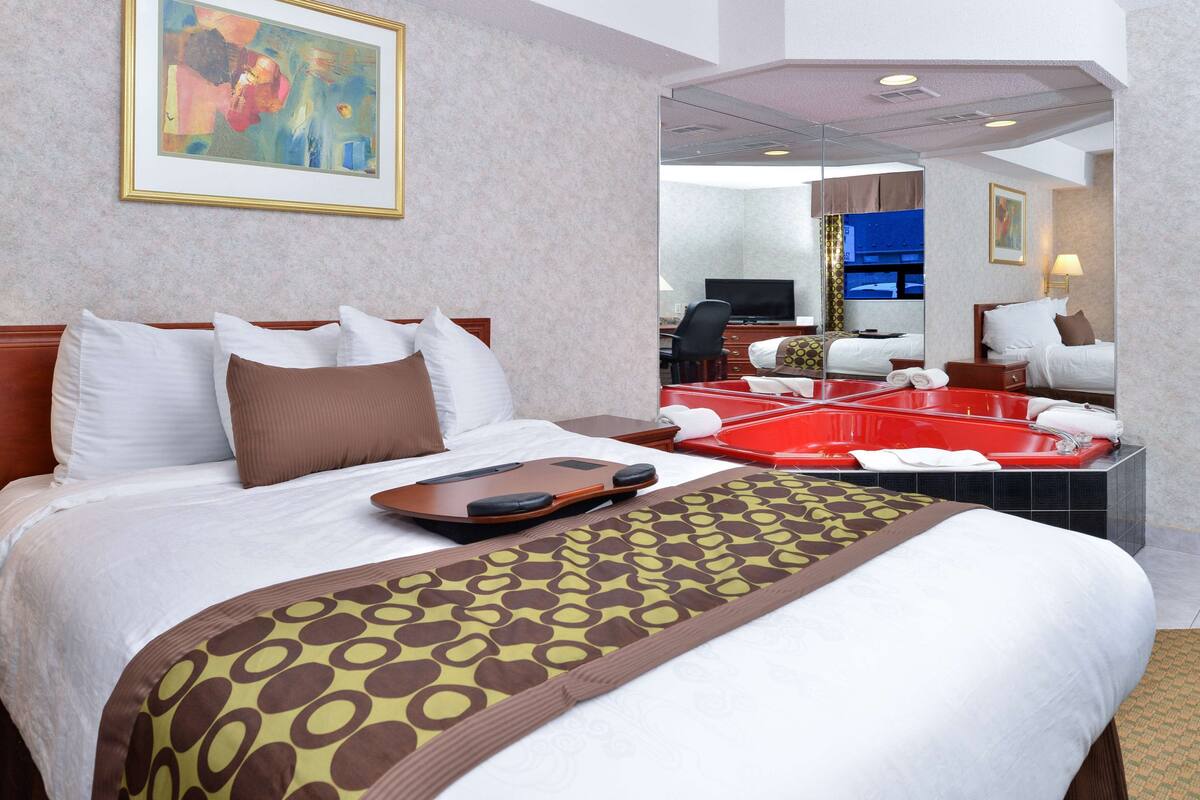 Quarto de hotel com cama grande, uma banheira vermelha, escrivaninha com computador e espelho refletindo o ambiente.