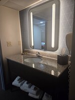 Uma casa de banho moderna com lavatório de granito, um espelho grande com iluminação integrada, um porta-toalhas e um dispensador de lenços de papel.