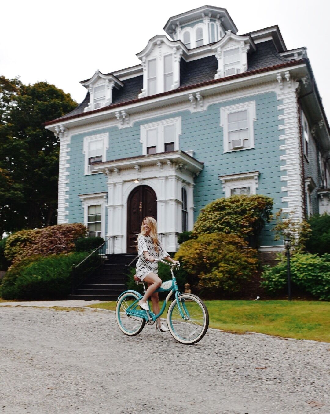 Uma mulher em uma bicicleta azul-turquesa em frente a um prédio grande, azul claro, com detalhes brancos e telhado escuro.