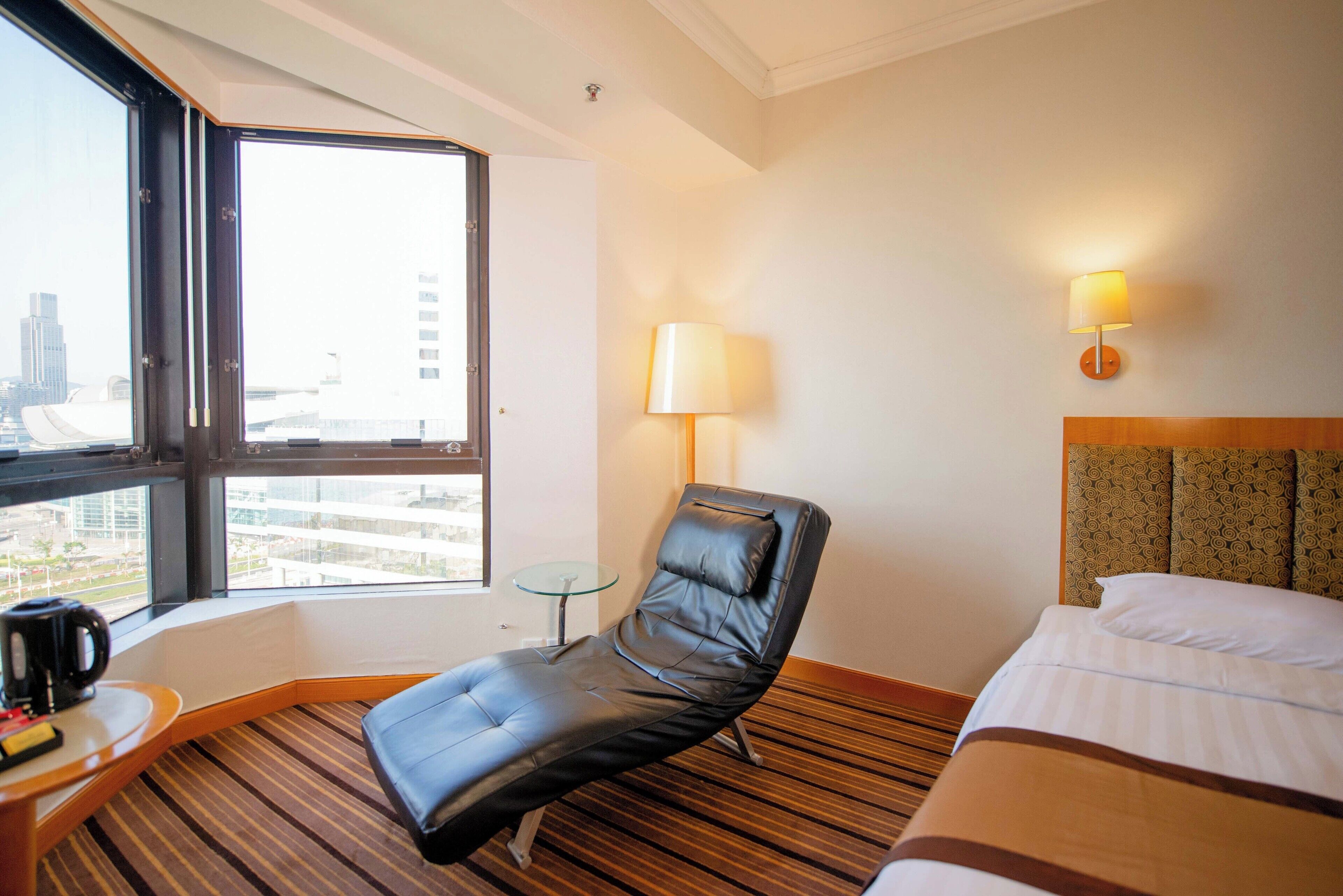 Quarto de hotel com uma janela grande, uma poltrona de couro, uma cama, uma mesa de cabeceira com abajur e vista para a paisagem urbana.