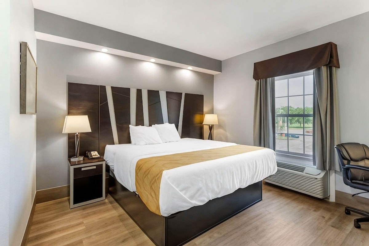 Quarto de hotel com cama, luminárias de cabeceira, uma cadeira, janela com cortinas e uma obra de arte na parede.