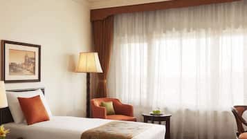 Quarto de hotel com cama, cadeira, mesa com abajur e uma janela grande com cortinas.