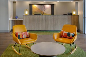 Uma área de recepção com design moderno, com um balcão de atendimento, duas poltronas de balanço com almofadas coloridas e uma mesa redonda.