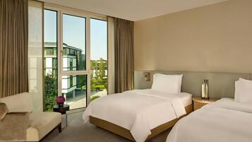 Quarto de hotel com duas camas, um sofá, uma mesa de cabeceira e uma grande janela com vista para um edifício e vegetação.