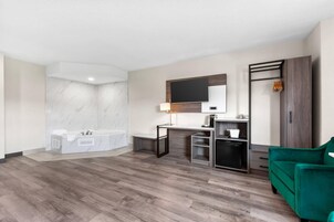 Quarto de hotel moderno com piso de madeira, sofá verde, televisão de tela plana, escrivaninha com luminária, frigobar e um espelho grande.
