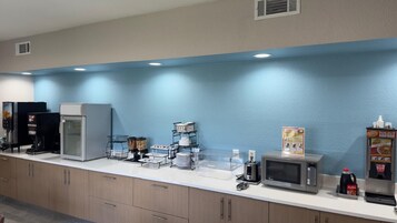 Espaço de café da manhã com serviço de buffet, máquinas de café, micro-ondas e dispensador de bebidas.