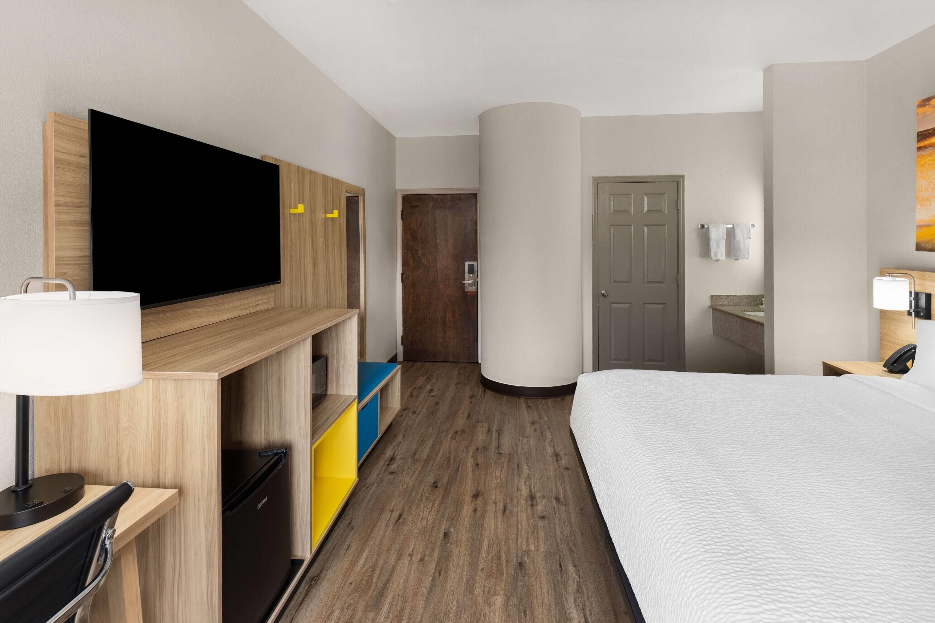 Quarto de hotel com cama, televisão, escrivaninha com luminária, minibar e banheiro com vaso sanitário e chuveiro.