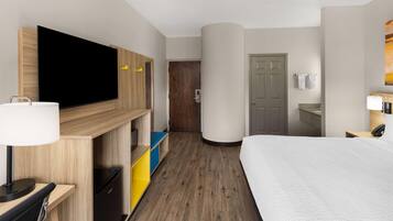 Quarto de hotel com cama, televisão, escrivaninha com luminária, minibar e banheiro com vaso sanitário e chuveiro.
