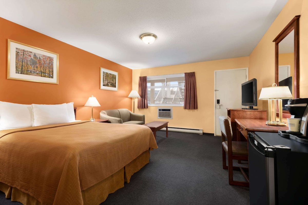 Quarto de hotel com uma cama grande, uma mesa com computador, uma cadeira, uma televisão e uma janela com cortinas.