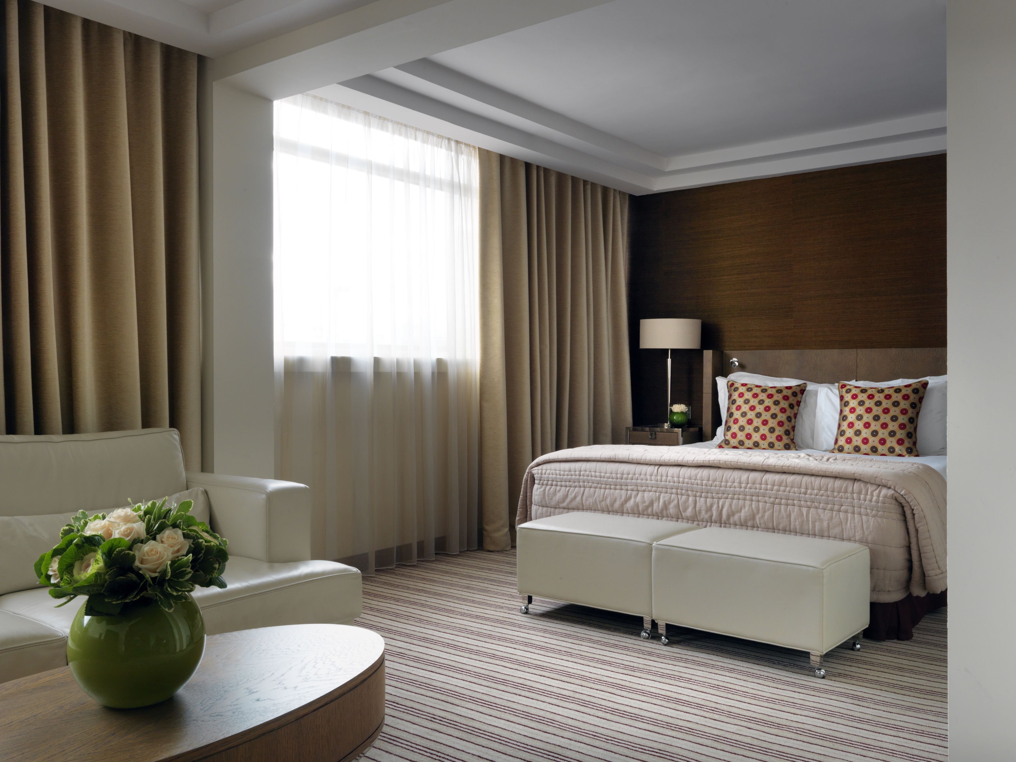 Quarto de hotel com cama, sofá, mesa de centro com um vaso de flores e janela com cortinas.