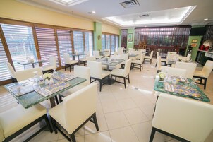 Um restaurante com várias mesas e cadeiras, grandes janelas com persianas e piso de azulejos.