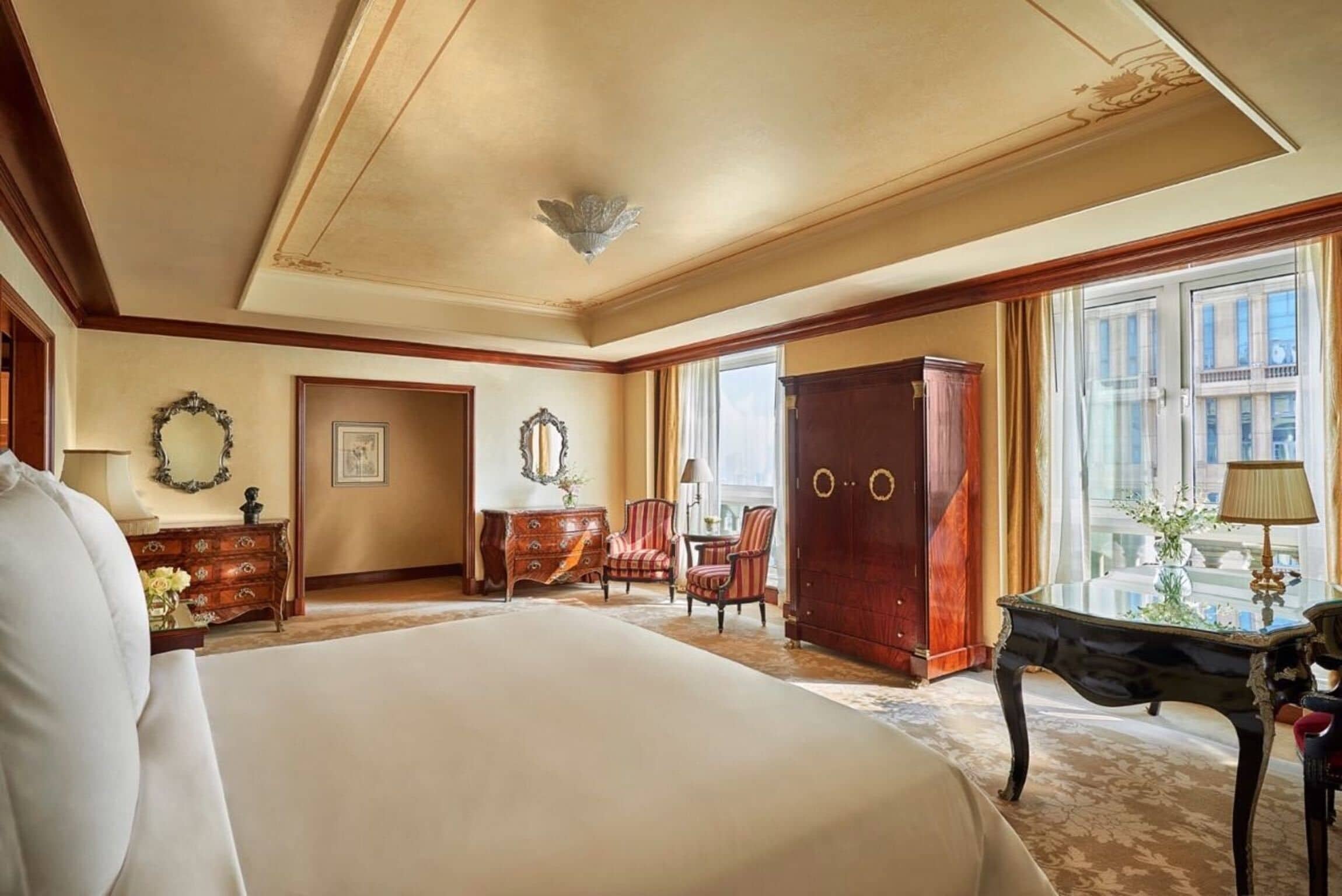Quarto de hotel espaçoso com uma cama grande, um guarda-roupa de madeira, uma escrivaninha com uma lâmpada e vista da cidade através de grandes janelas.