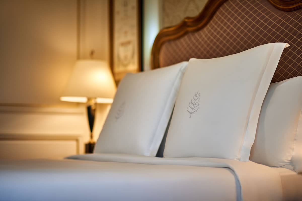 Cama bem arrumada com lençóis brancos e almofadas decorativas, abajur e cabeceira de madeira.