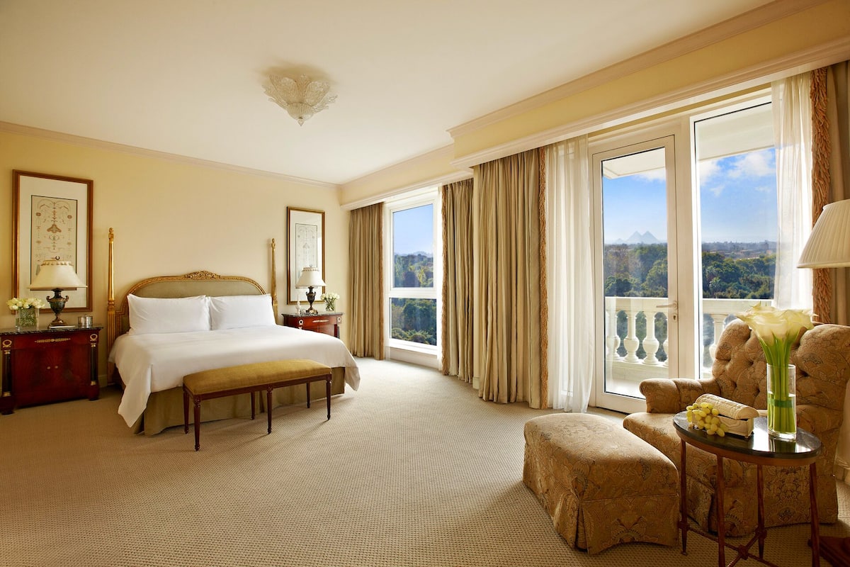 Quarto de hotel com uma cama grande, um espaço de estar com uma poltrona e mesa, vista para as montanhas pela janela e um lustre.