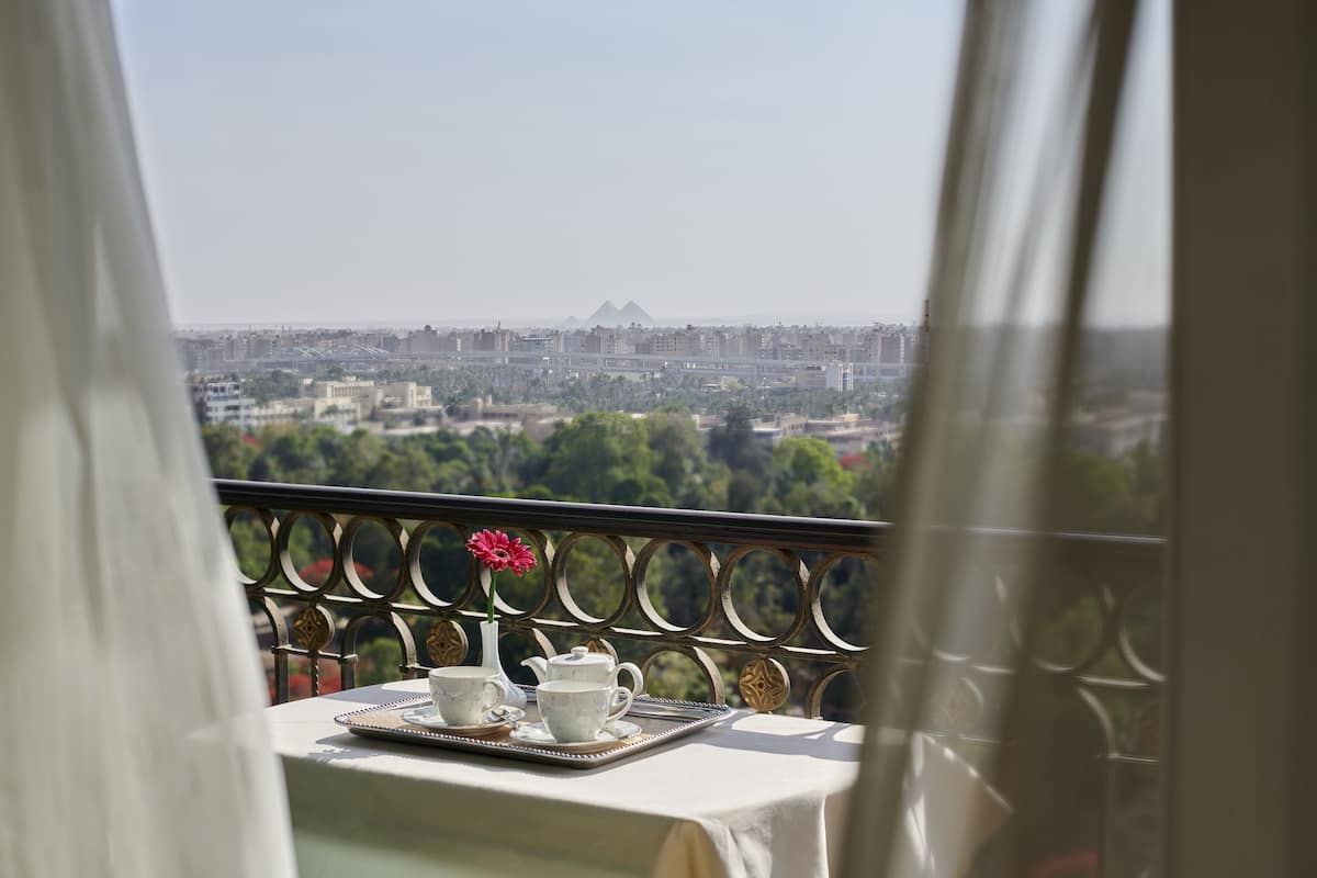 Uma varanda com uma mesa posta para chá, com vista para uma paisagem urbana e uma pirâmide imponente ao longe.