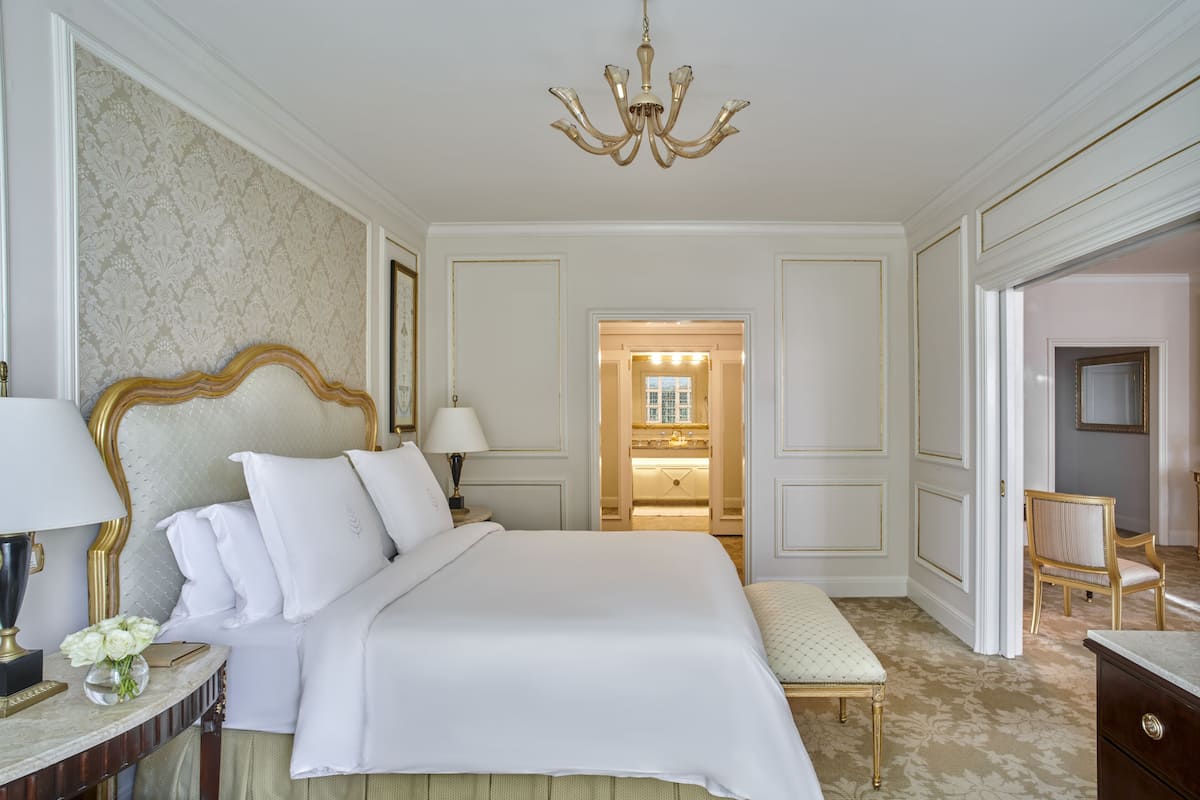 Um quarto espaçoso com uma cama grande, uma mesa de cabeceira com abajur, um banco e uma cadeira.