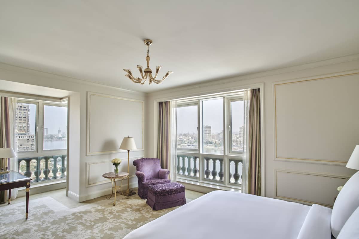 Quarto de hotel com uma cama grande, uma poltrona roxa, uma mesa pequena e um lustre.