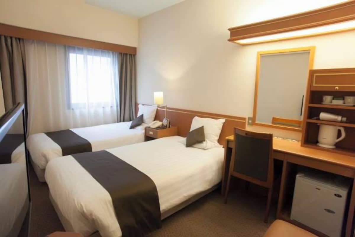 Quarto de hotel com duas camas, uma escrivaninha, uma cadeira, um pequeno frigorífico e um espelho.