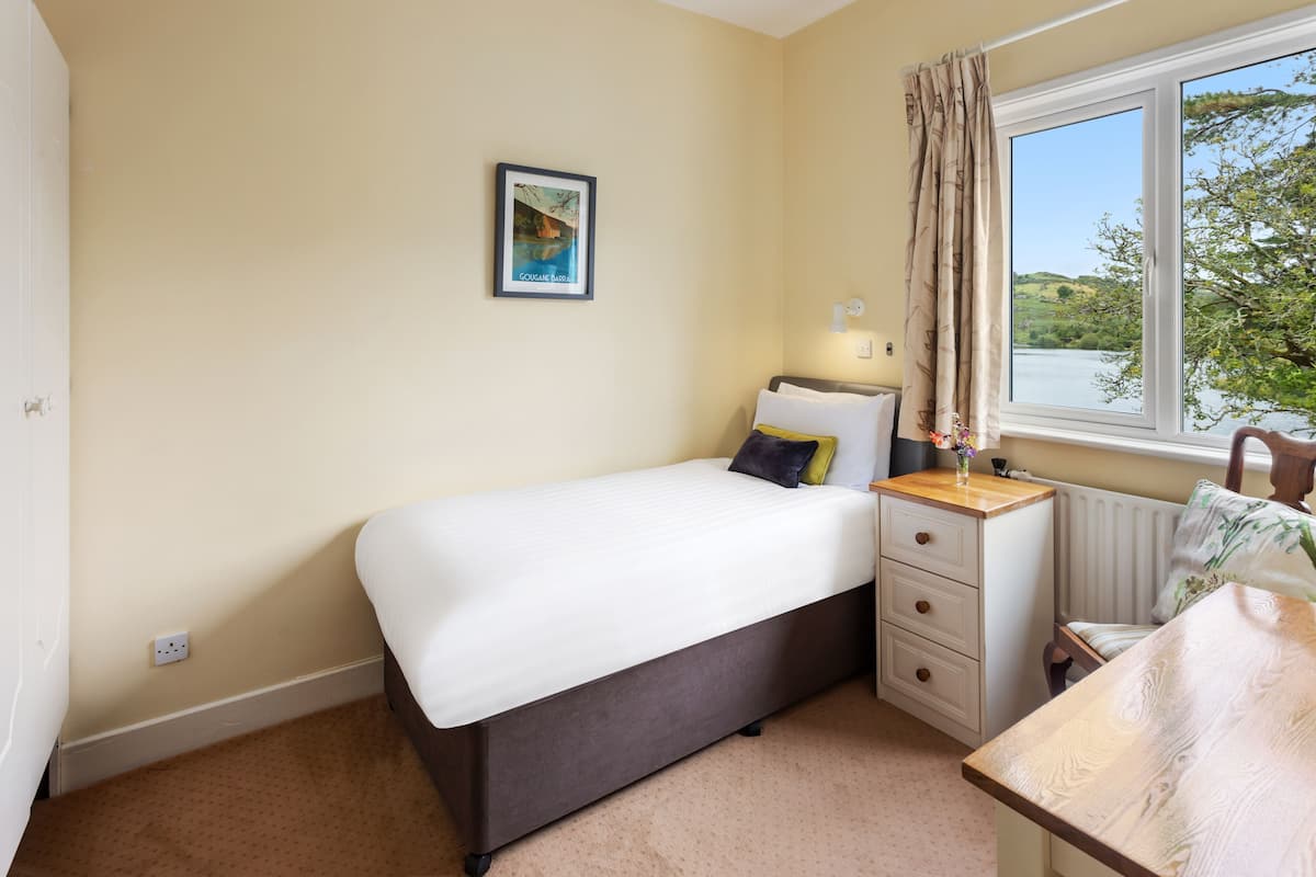 Quarto com cama, mesa de cabeceira com abajur, janela com vista para o verde e um quadro na parede.