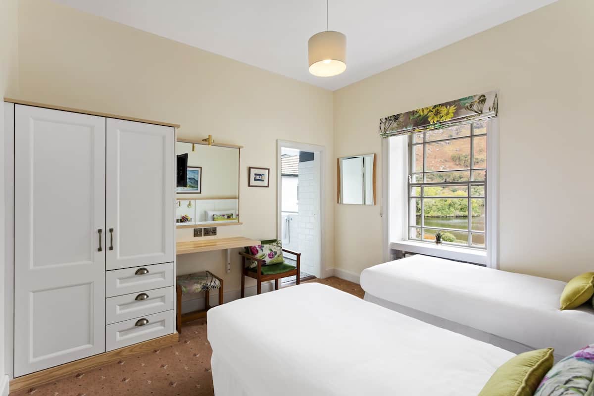 Quarto com duas camas de solteiro, uma escrivaninha com cadeira, guarda-roupa e janela com vista para área verde.