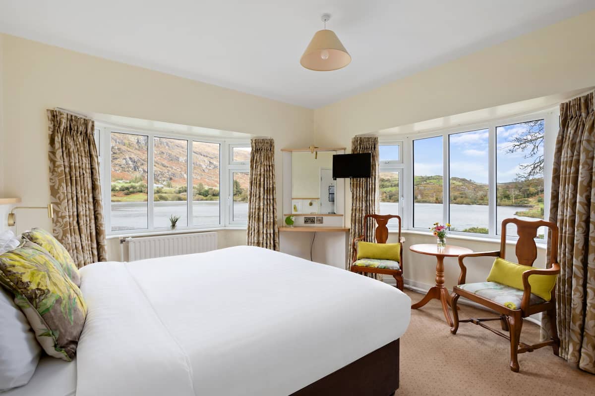 Quarto de hotel com cama grande, duas cadeiras, uma mesinha, televisão e vista para um lago.
