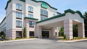 Um hotel com vários andares, fachada verde e bege, entrada destacada com galeria coberta e placa com a inscrição "Wingate by Wyndham".