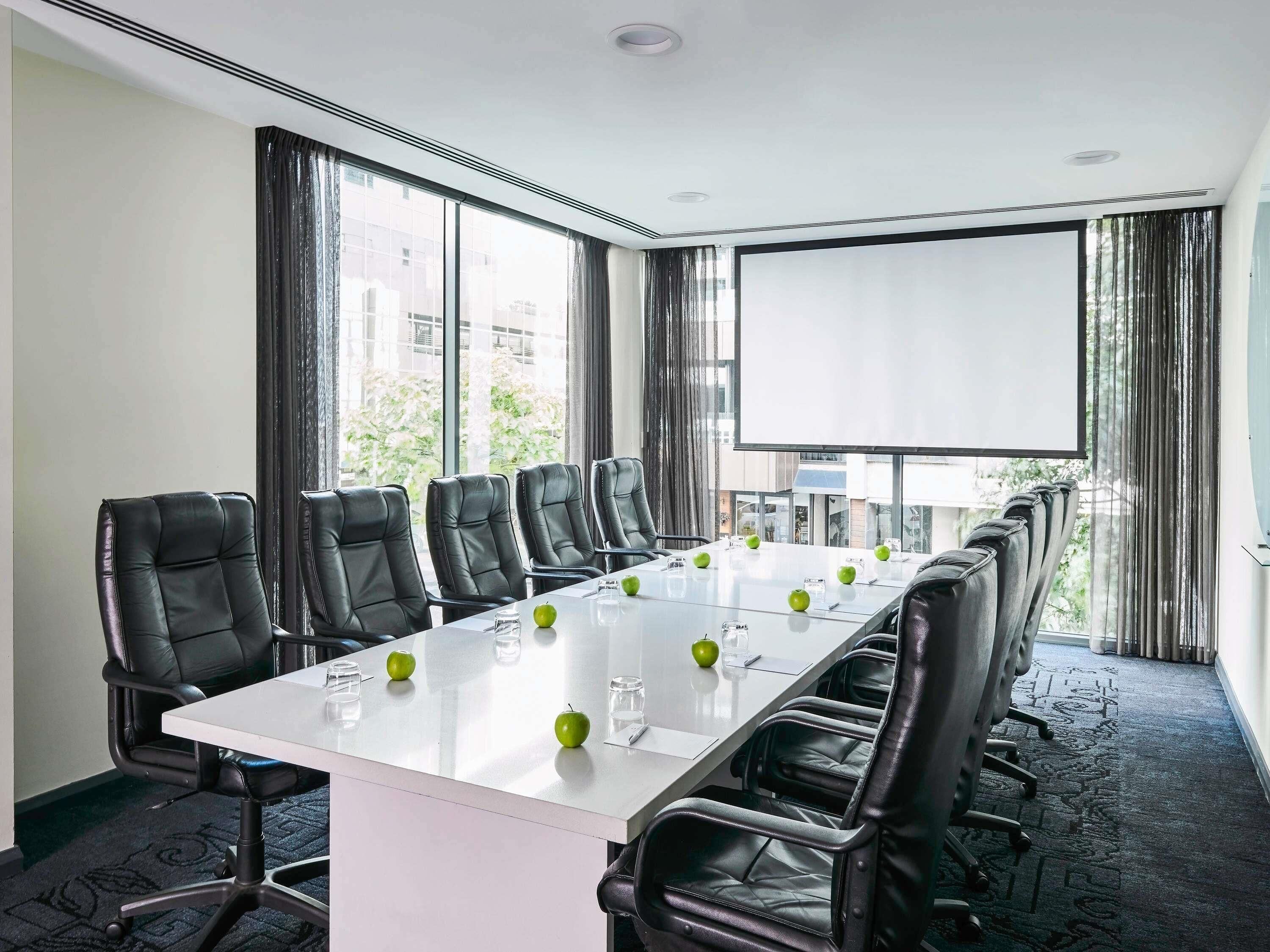 Uma sala de conferência com uma mesa comprida, cadeiras de couro preto, uma tela de projetor e janelas amplas.