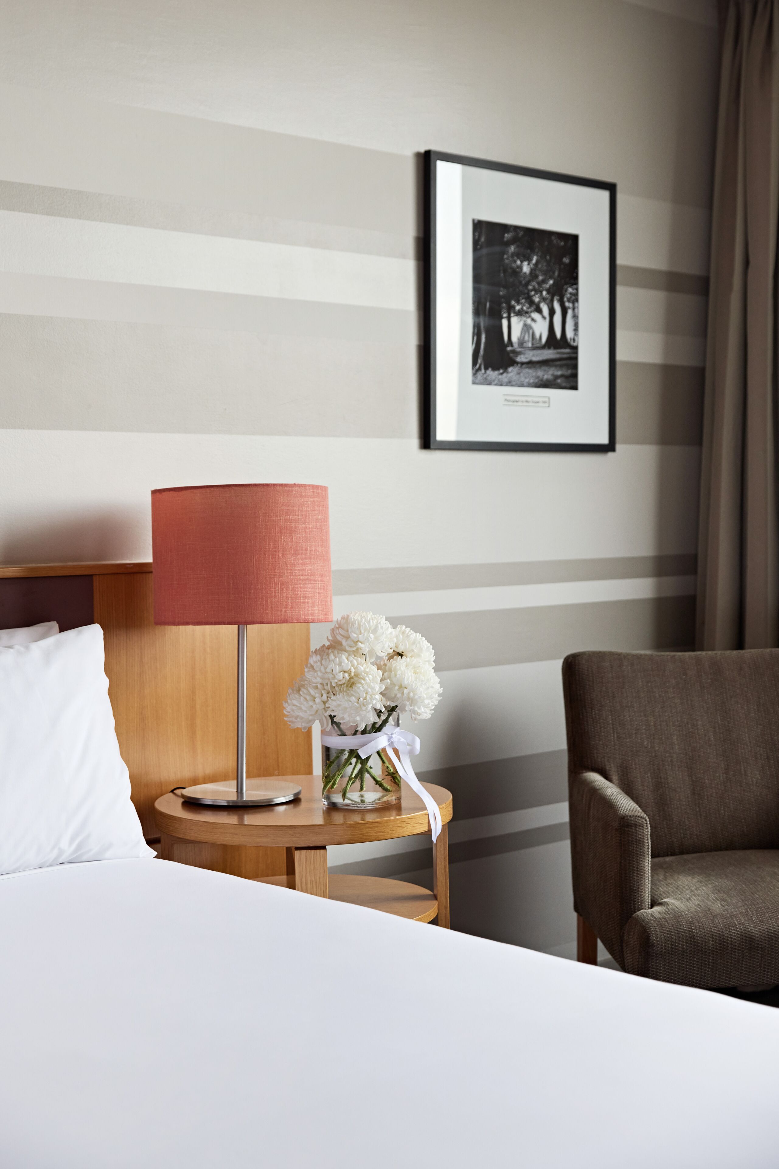 Quarto de hotel com uma cama, uma mesa redonda com uma luminária e flores, um quadro na parede e uma cadeira.