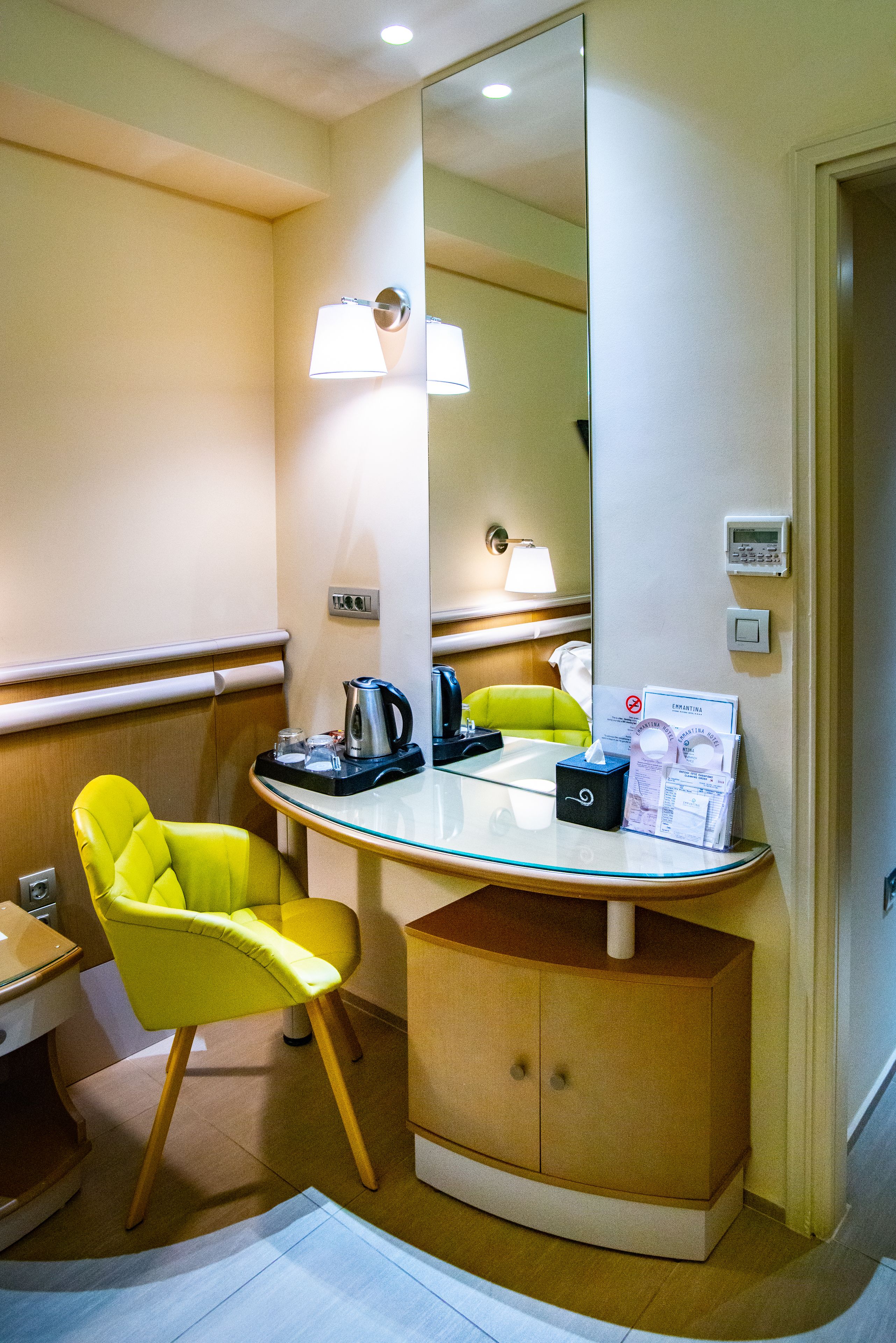 Quarto de hotel com uma cadeira amarela, uma mesa de vidro, um armário, um espelho e um abajur.