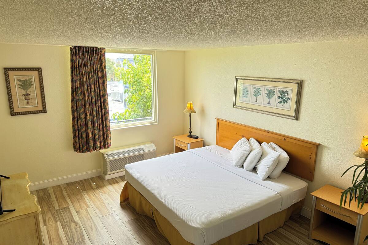 Quarto de hotel com cama, mesa de cabeceira, luminária, janela com cortinas e quadros nas paredes.