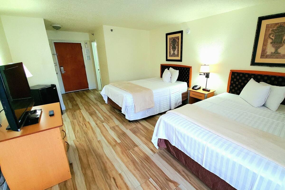 Quarto de hotel com duas camas, uma mesa com um computador e uma porta que dá para outro cômodo.