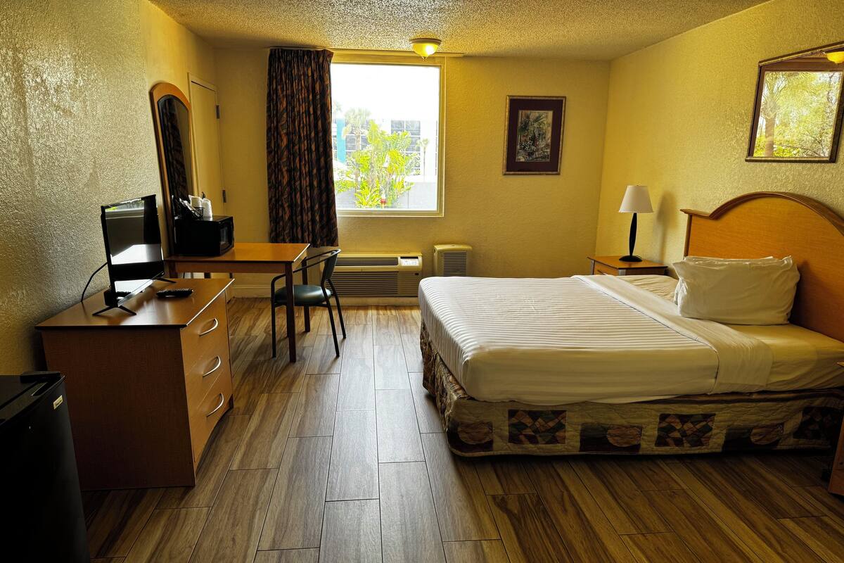 Quarto de hotel com cama, escrivaninha, cadeira, televisão e janela com vista para árvores.