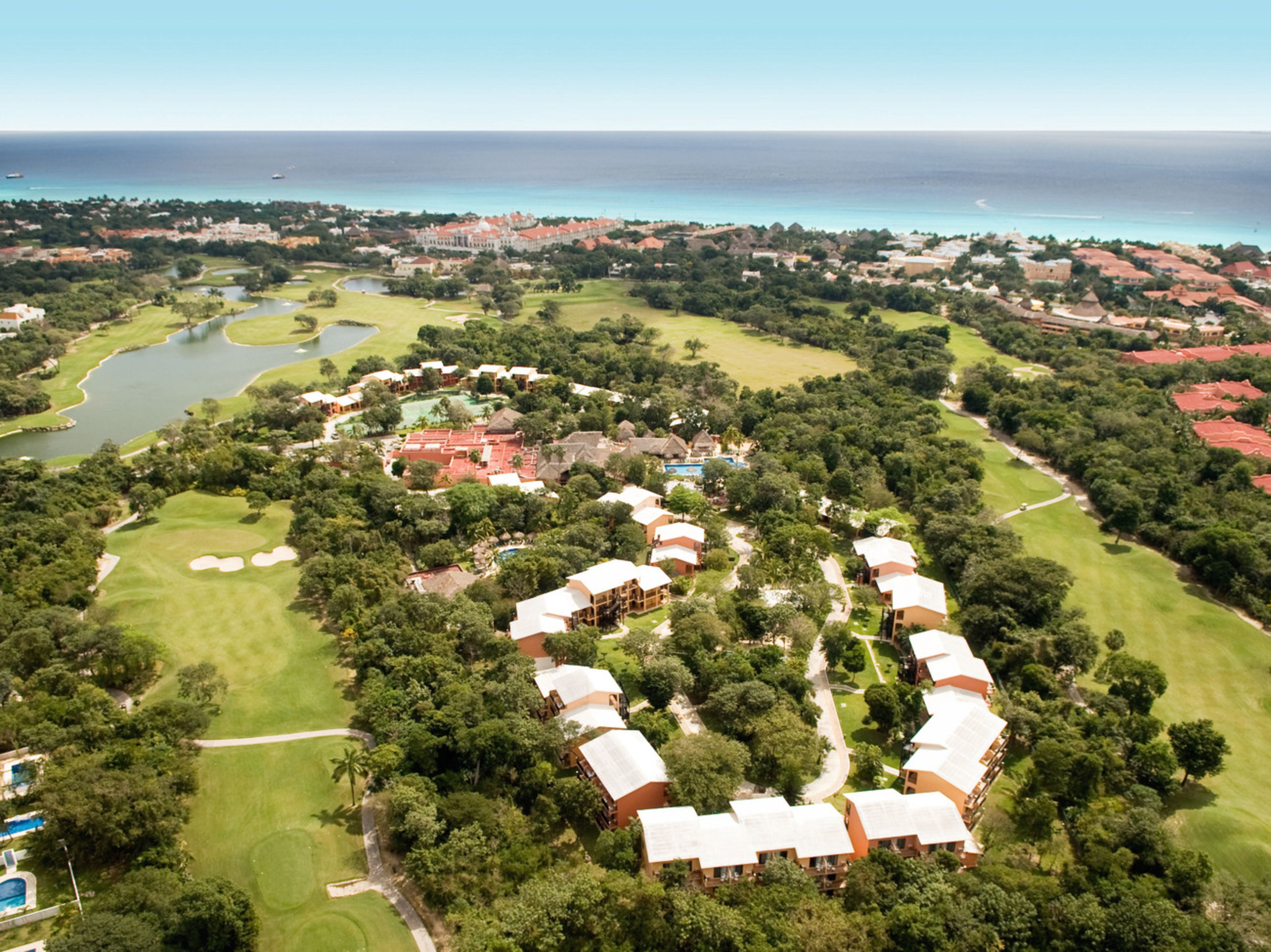 Vista aérea de um resort com vários edifícios dispostos em formato de semicírculo, rodeado por vegetação exuberante e um campo de golfe.