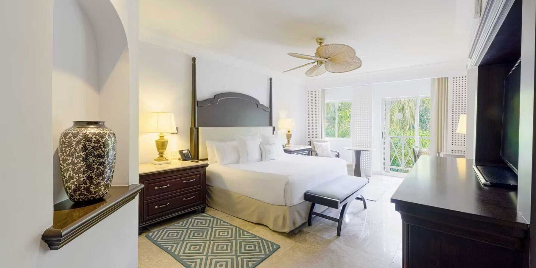 Quarto de hotel com cama, mesas de cabeceira, escrivaninha, cadeira, televisão, ventoinha no teto e janela com vista.