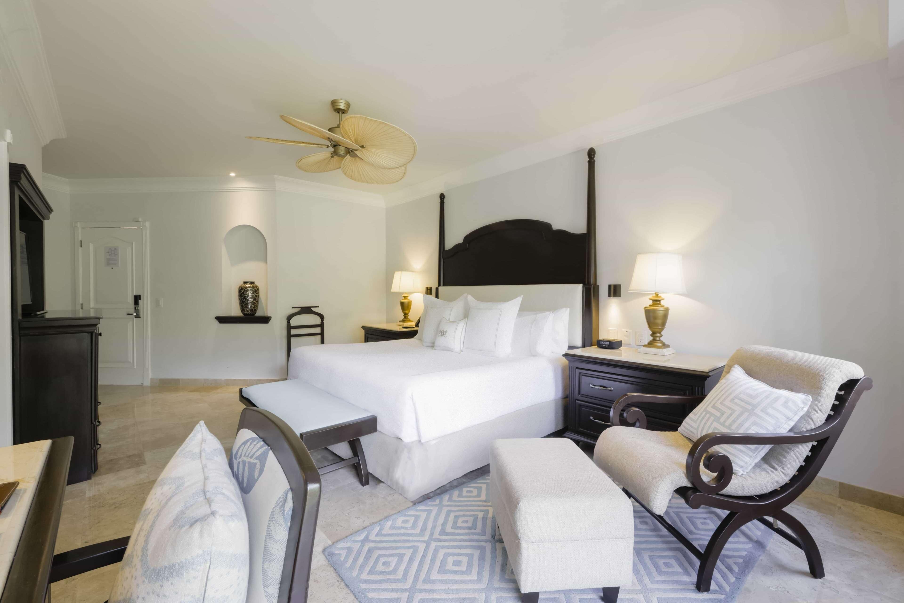 Quarto de hotel com cama, mesas de cabeceira, escrivaninha, cadeira, ventilador de teto e tapete estampado.