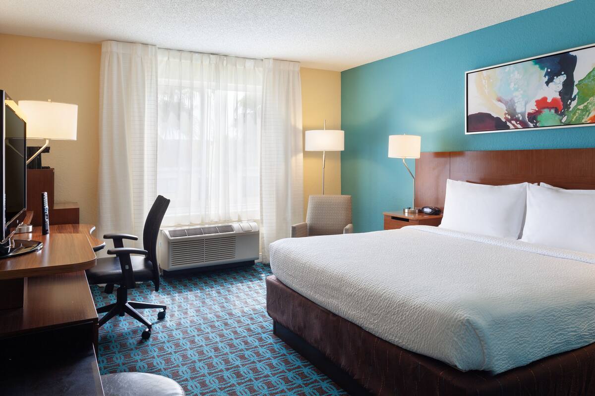 Quarto de hotel com uma cama grande, escrivaninha com computador, ar condicionado e janela com cortinas.