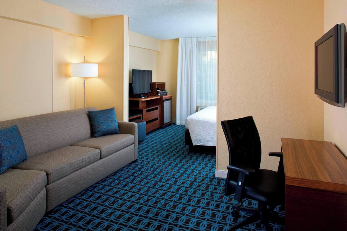 Quarto de hotel com sofá, cama, escrivaninha, cadeira, televisão e luminária.