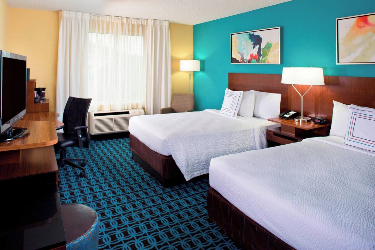 Quarto de hotel com duas camas, uma secretária com televisão, uma cadeira e um tapete estampado.