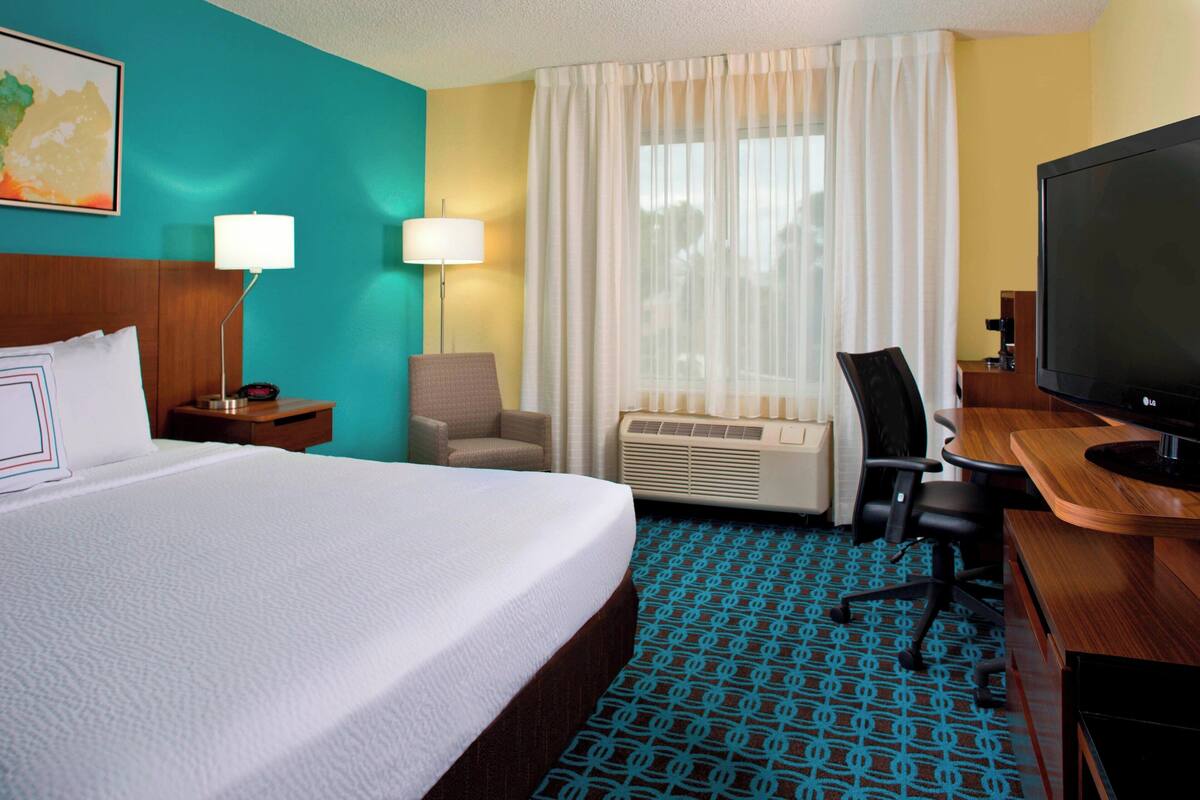 Quarto de hotel com uma cama grande, uma escrivaninha com computador, uma cadeira, uma televisão e um ar condicionado.