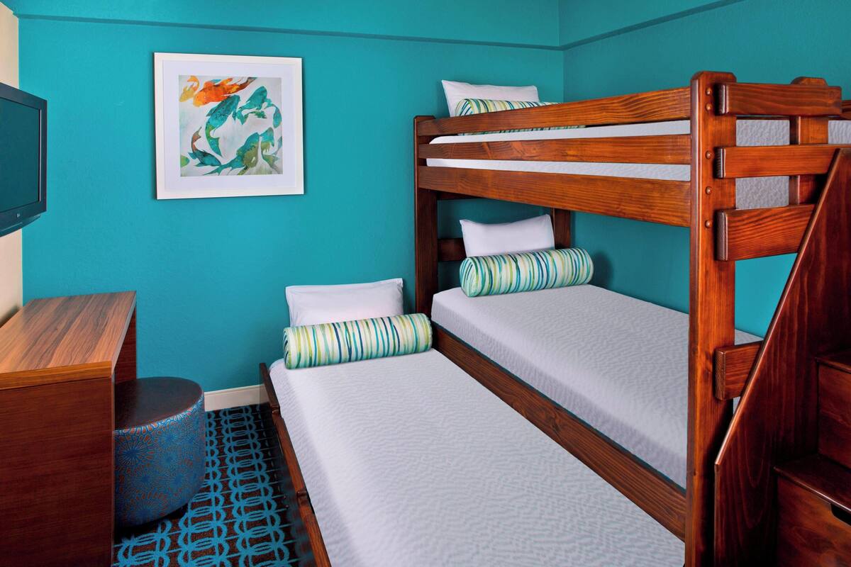 Quarto com beliche de madeira, uma escrivaninha pequena, uma cadeira e um tapete estampado.