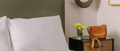 Cama bem arrumada com lençóis brancos, uma mesinha de cabeceira com um vaso de flores amarelas e uma bolsa de couro marrom sobre a mesa.