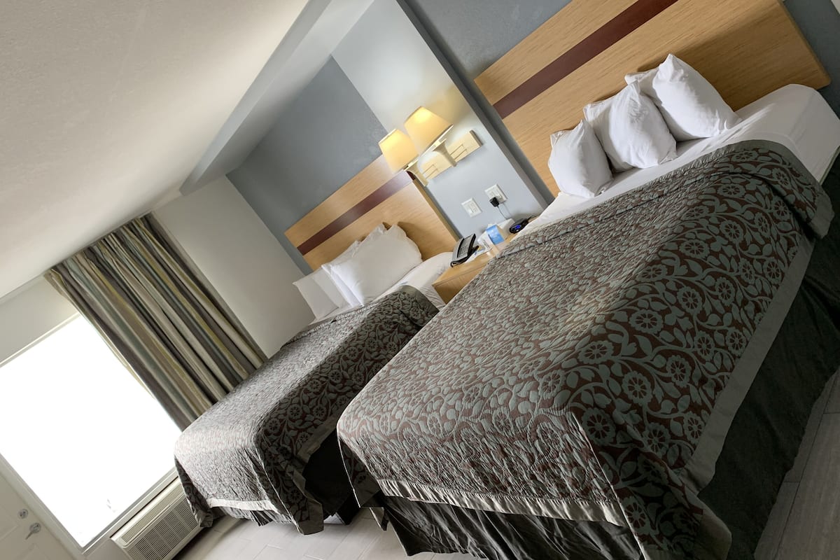 Quarto de hotel com duas camas, uma escrivaninha, uma luminária e uma janela com cortinas.