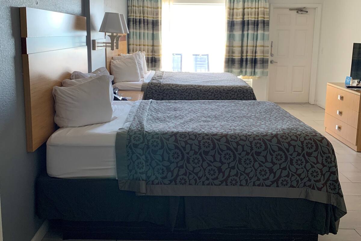 Quarto de hotel com duas camas, cabeceira de madeira, mesa de cabeceira com luminária e vista para o exterior através de uma janela com cortinas.