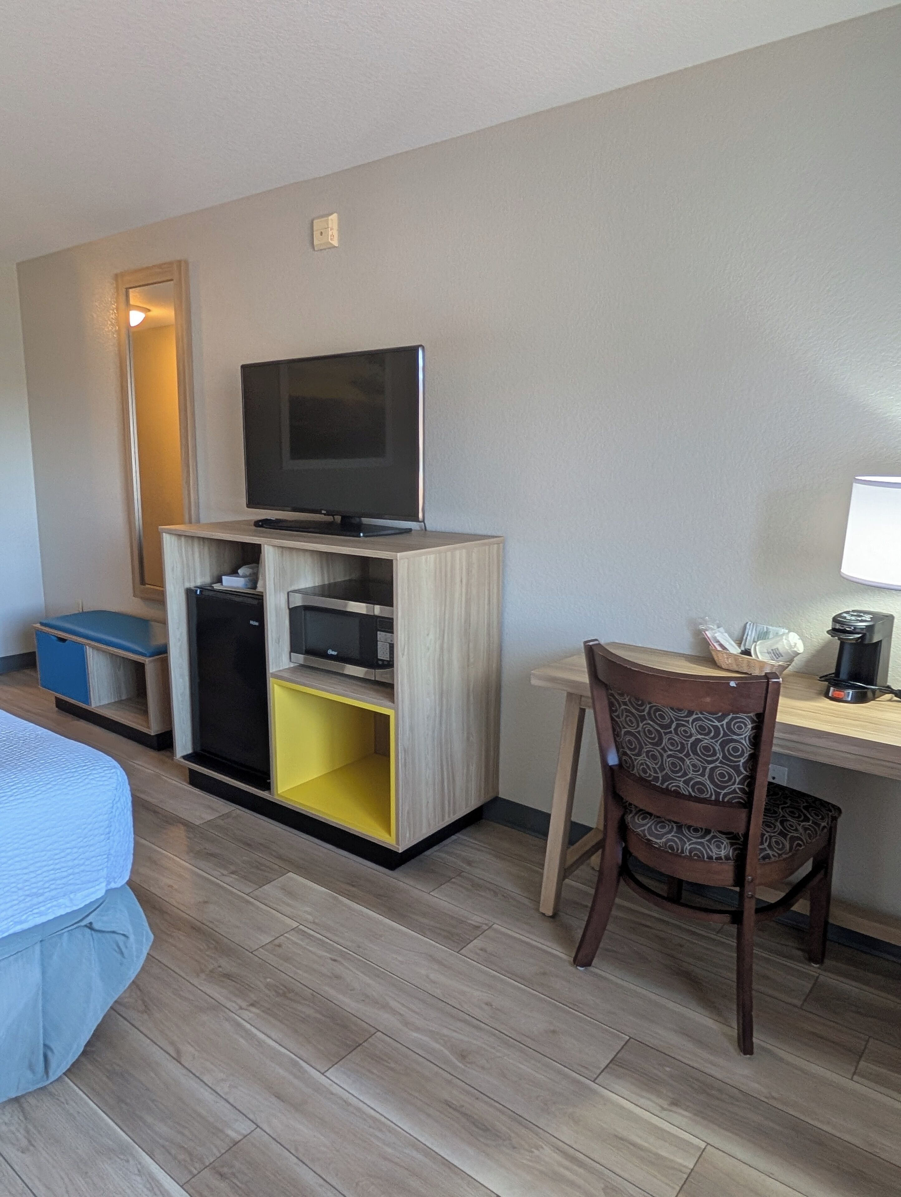 Quarto de hotel com cama, escrivaninha com cadeira, televisão, micro-ondas e cafeteira.